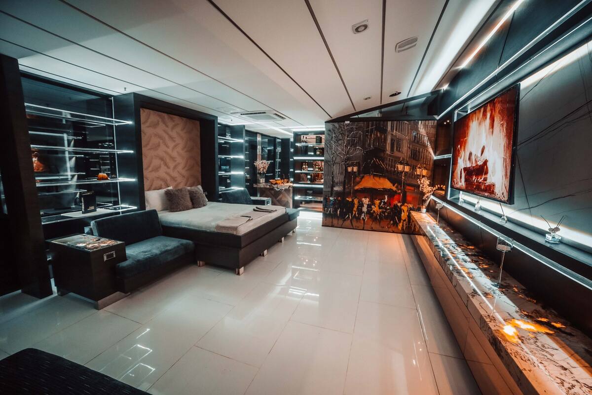 Quarto de hotel moderno com cama, área de estar, televisão grande e parede decorativa.