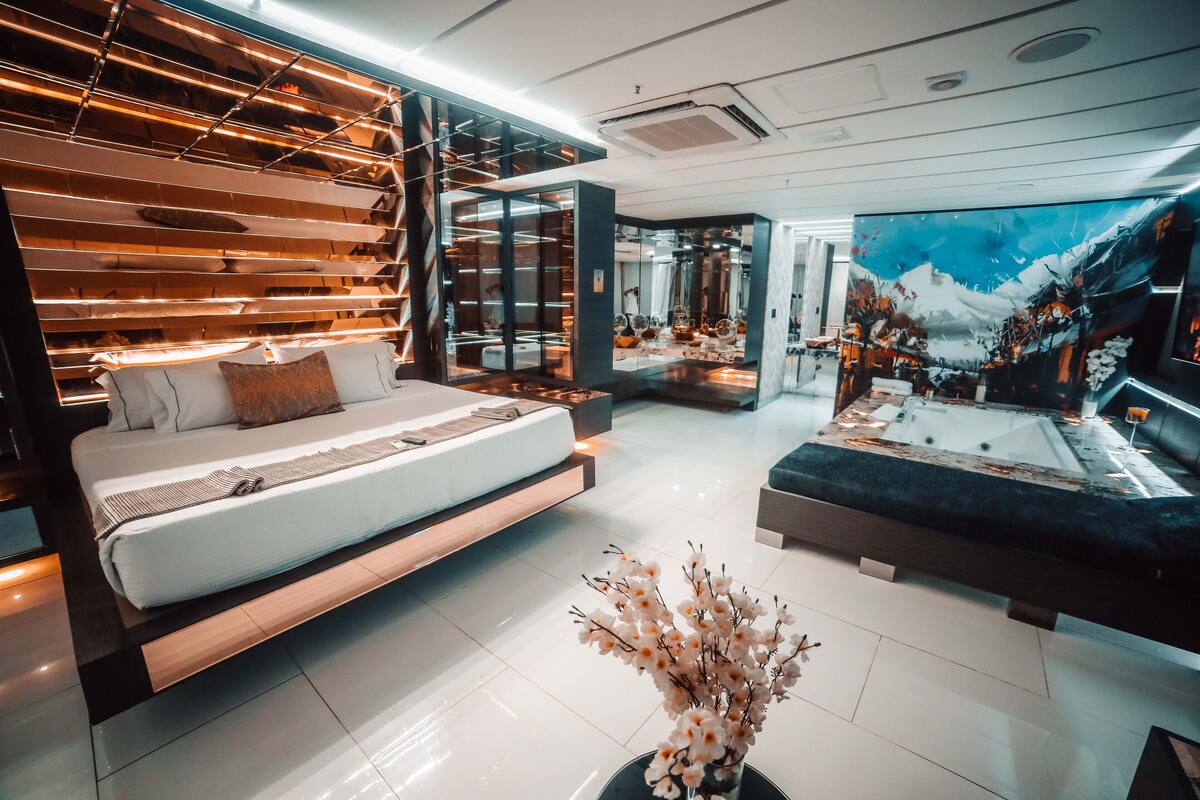 Quarto de hotel moderno com uma cama grande, área de estar, TV e uma planta decorativa.