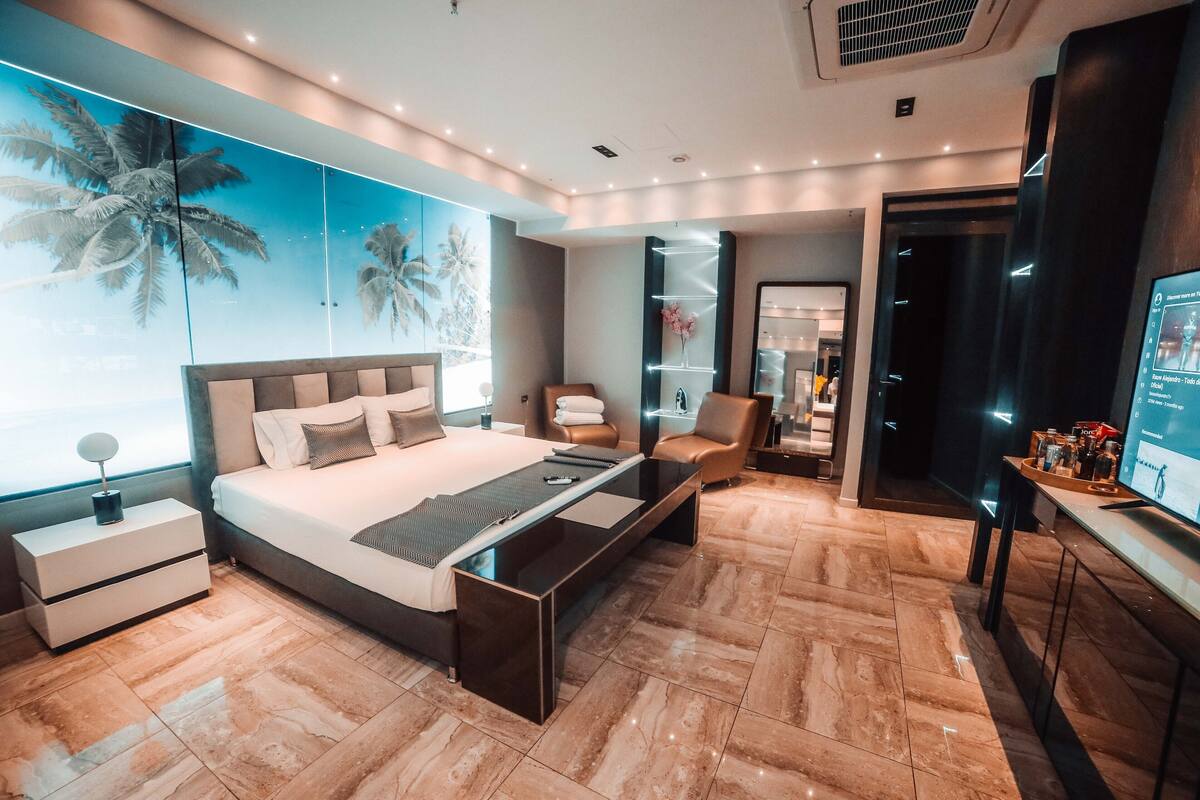 Quarto moderno com cama, mesas de cabeceira, escrivaninha, televisão e uma janela grande com arte de palmeiras.