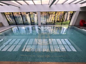 Piscina interior com água cristalina, rodeada por amplas janelas com vista para uma paisagem arborizada.