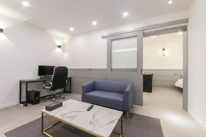 Quarto de hotel moderno com uma mesa de centro de mármore, um sofá azul, uma escrivaninha com computador e um espelho grande.