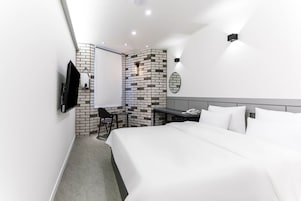 Quarto de hotel moderno com uma cama grande, uma escrivaninha com cadeira, televisão de tela plana na parede e um pequeno espaço de estar com uma poltrona acolchoada e um quadro branco.