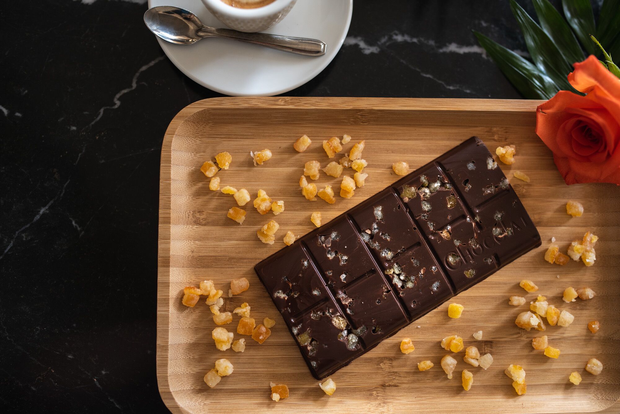 Uma barra de chocolate sobre tábua de madeira com pedaços de caramelo, uma colher, uma chávena e uma rosa em superfície escura.