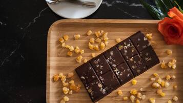 Uma barra de chocolate sobre tábua de madeira com pedaços de caramelo, uma colher, uma chávena e uma rosa em superfície escura.