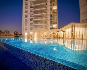 Uma piscina com borda azulejada, rodeada por um edifício moderno com várias varandas e janelas de vidro.