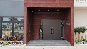Entrada de um edifício moderno, com uma porta grande, degraus de acesso e uma pequena área de jardim com plantas.