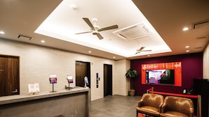 Uma área de recepção de hotel com design moderno, incluindo um balcão de atendimento, espaço para sentar e uma tela de televisão exibindo um videoclipe.
