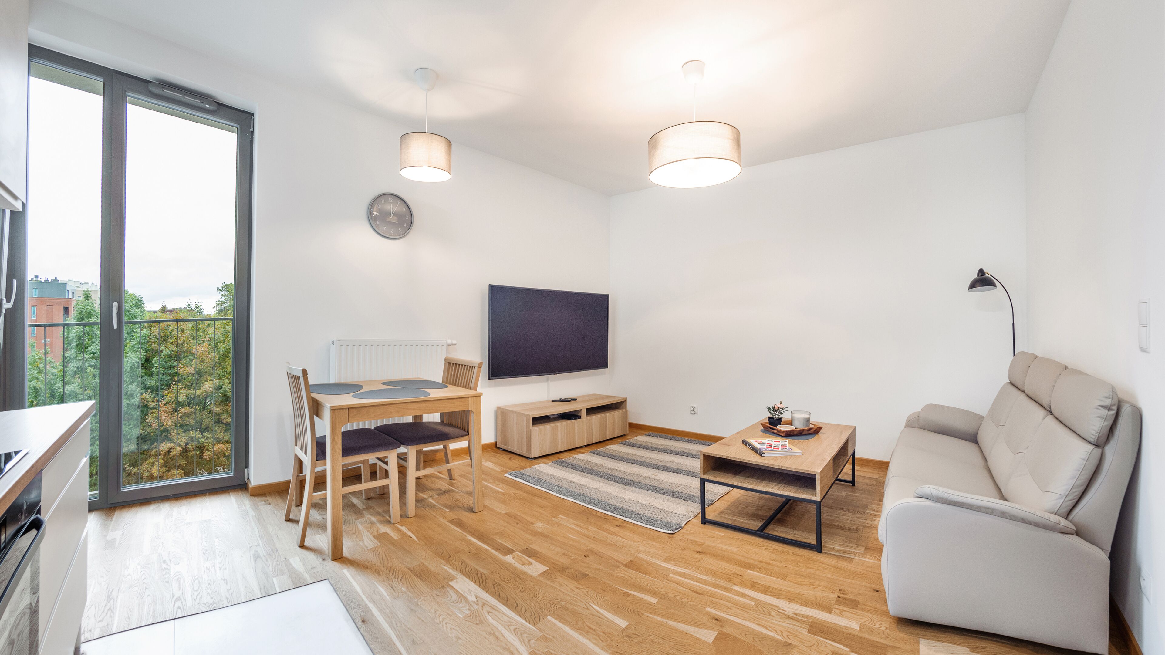 Uma sala de estar moderna com piso de madeira, TV de tela plana, sofá, mesa de jantar com cadeiras e uma grande janela com vista para árvores.