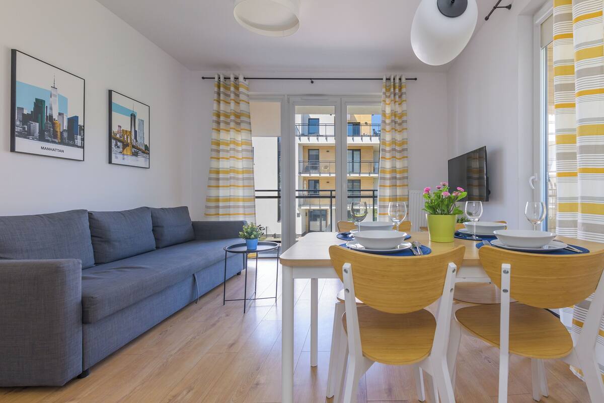Uma sala de estar moderna com um sofá cinzento, uma mesa de jantar com cadeiras e uma varanda com cortinas listradas de amarelo.