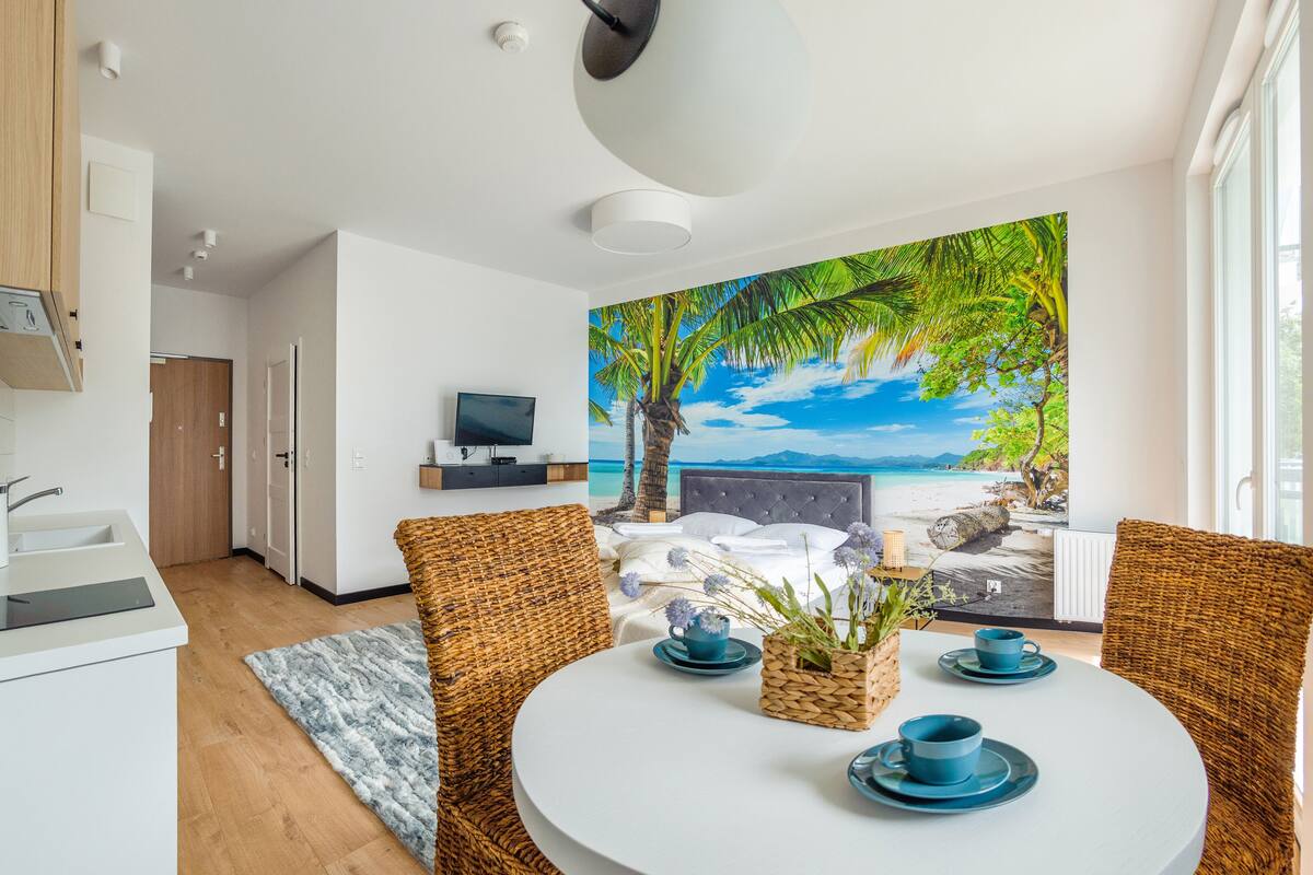 Uma sala de estar moderna com um mural de praia, uma mesa de jantar redonda com louças azuis, cadeiras de vime e uma cozinha com armários brancos e forno embutido.