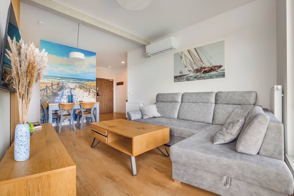Uma sala de estar moderna com um sofá cinzento, mesa de centro de madeira e cadeiras. Há um quadro com uma cena de praia na parede, e um aparelho de ar condicionado fixado acima do sofá.