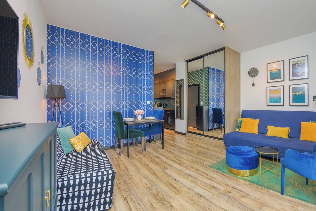 Uma sala de estar moderna com um sofá azul, um banquinho estampado e uma área de refeições com mesa e cadeiras.