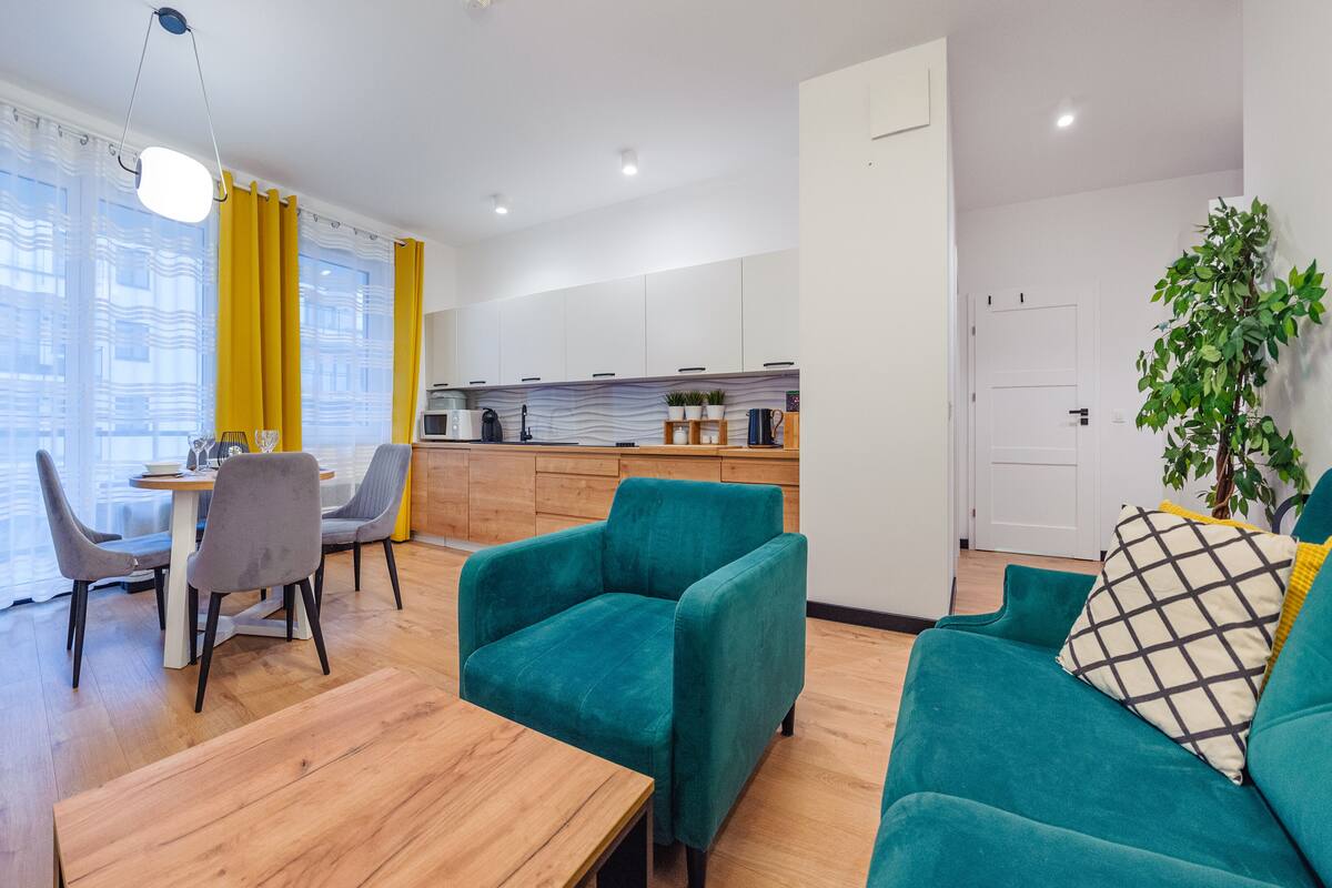 Uma sala de estar moderna com sofás azul-esverdeados, uma mesa de jantar de madeira e uma cozinha com armários brancos.