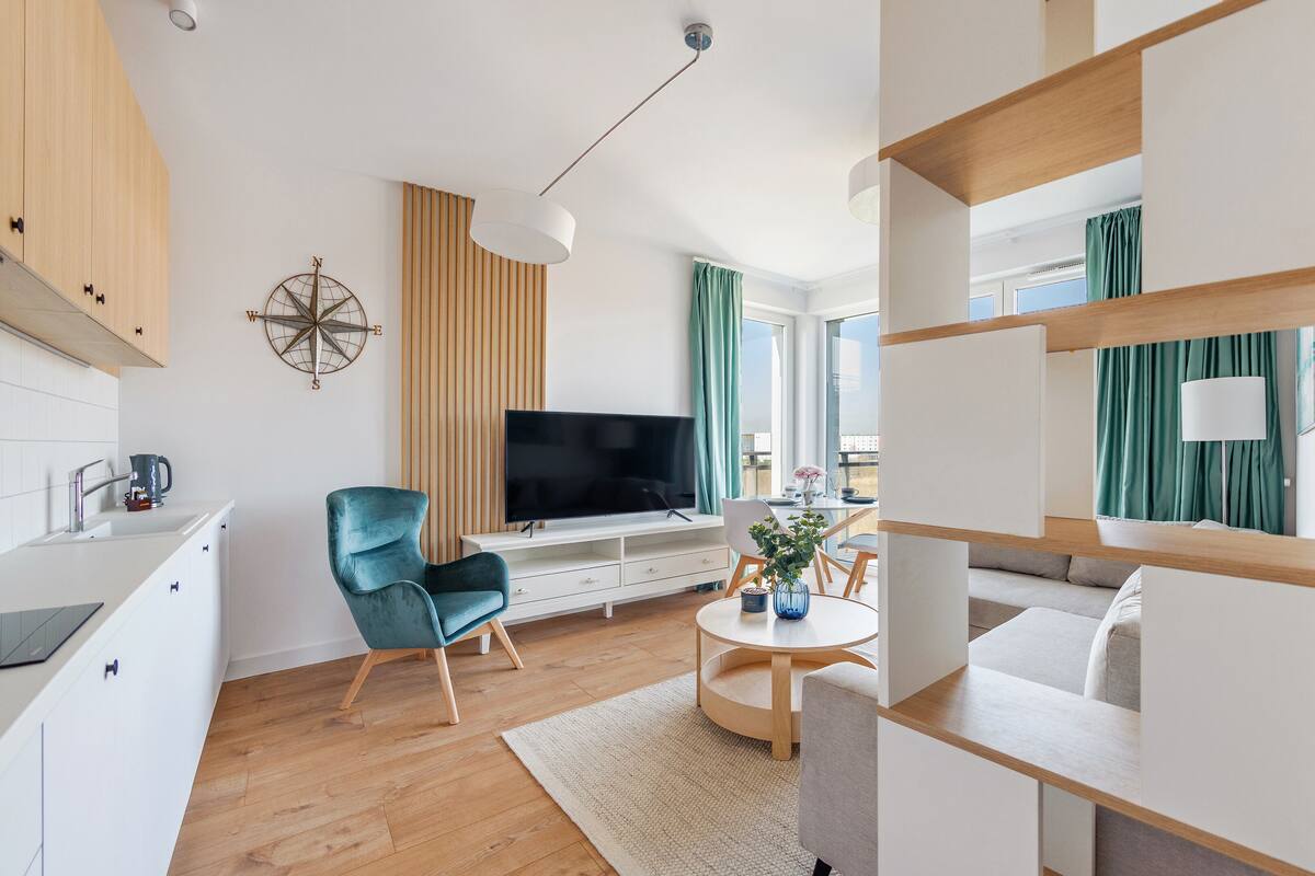 Uma sala de estar moderna com televisão, uma poltrona azul, uma mesa de centro de madeira e vista da cidade através de grandes janelas.