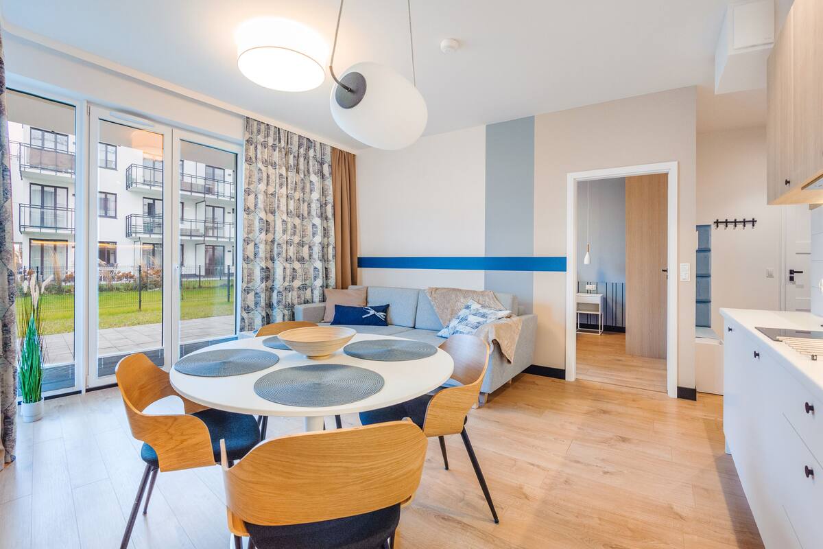 Uma sala de estar moderna com uma mesa de jantar redonda, cadeiras de madeira, um sofá e uma janela grande com vista para uma área residencial.