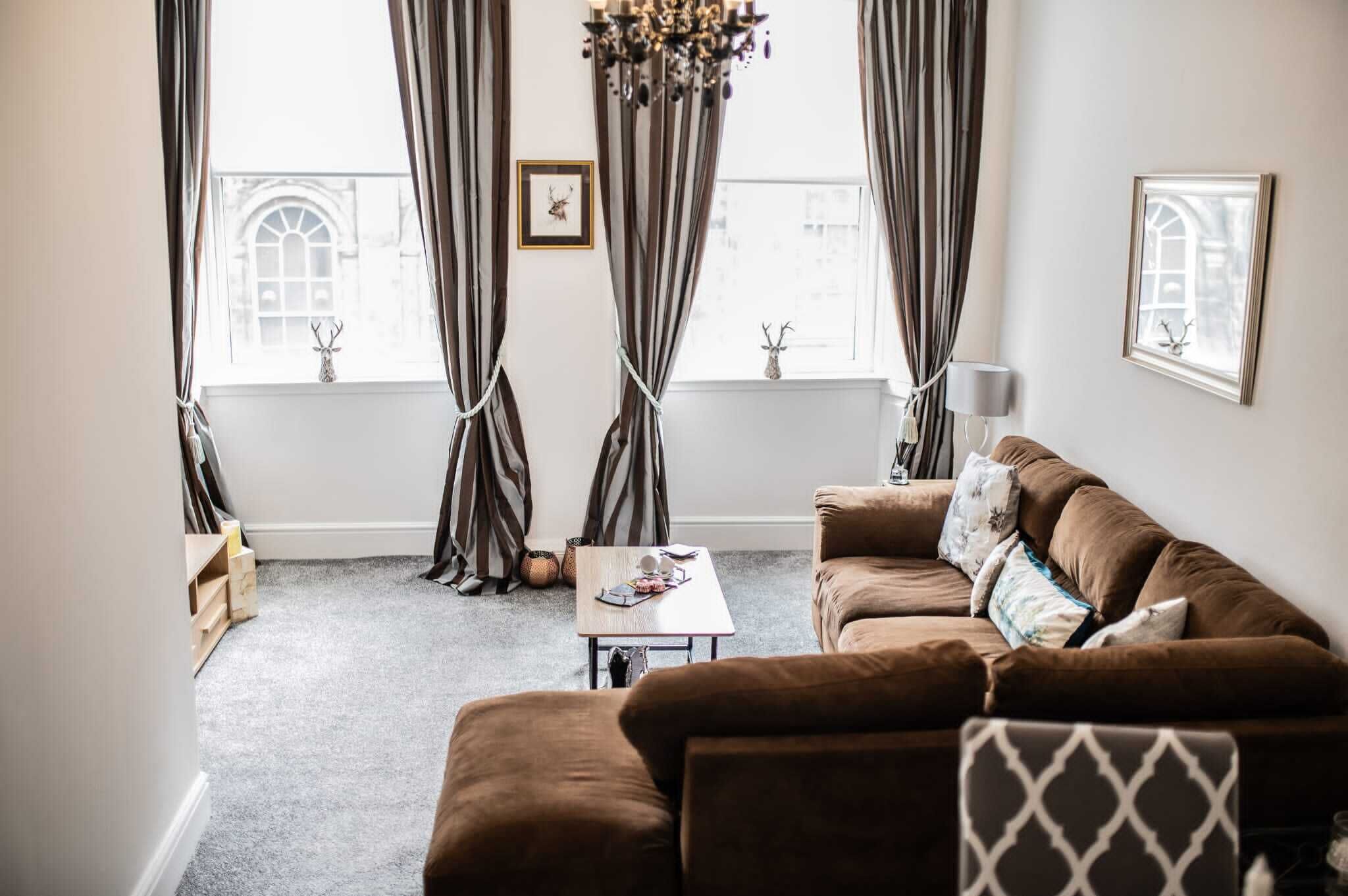 Uma sala de estar com um sofá marrom, uma mesa pequena e um candelabro. Há cortinas nas janelas e um quadro na parede.