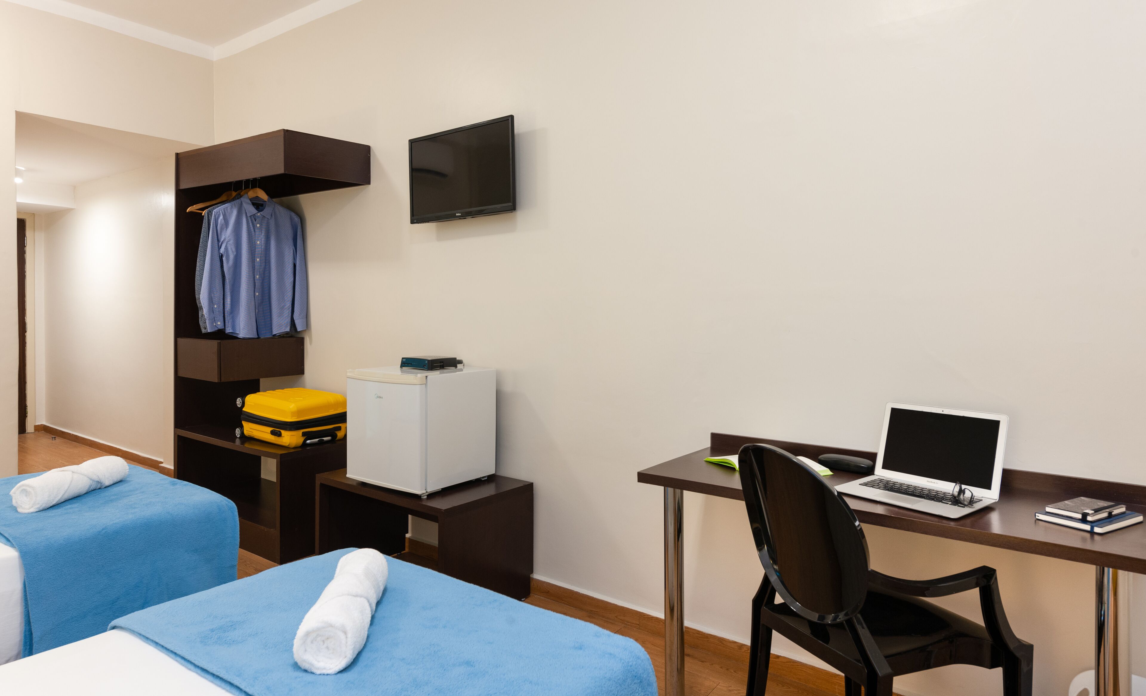 Quarto de hotel com duas camas, uma escrivaninha com um laptop, televisão e uma pequena geladeira.
