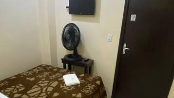 Quarto de hotel com cama, televisão na parede, porta e ventilador.