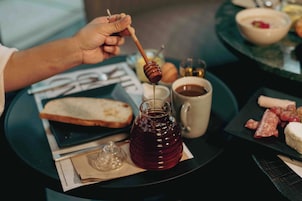 Uma bandeja com um copo de mel, uma xícara de café, fatias de pão e um prato de embutidos.