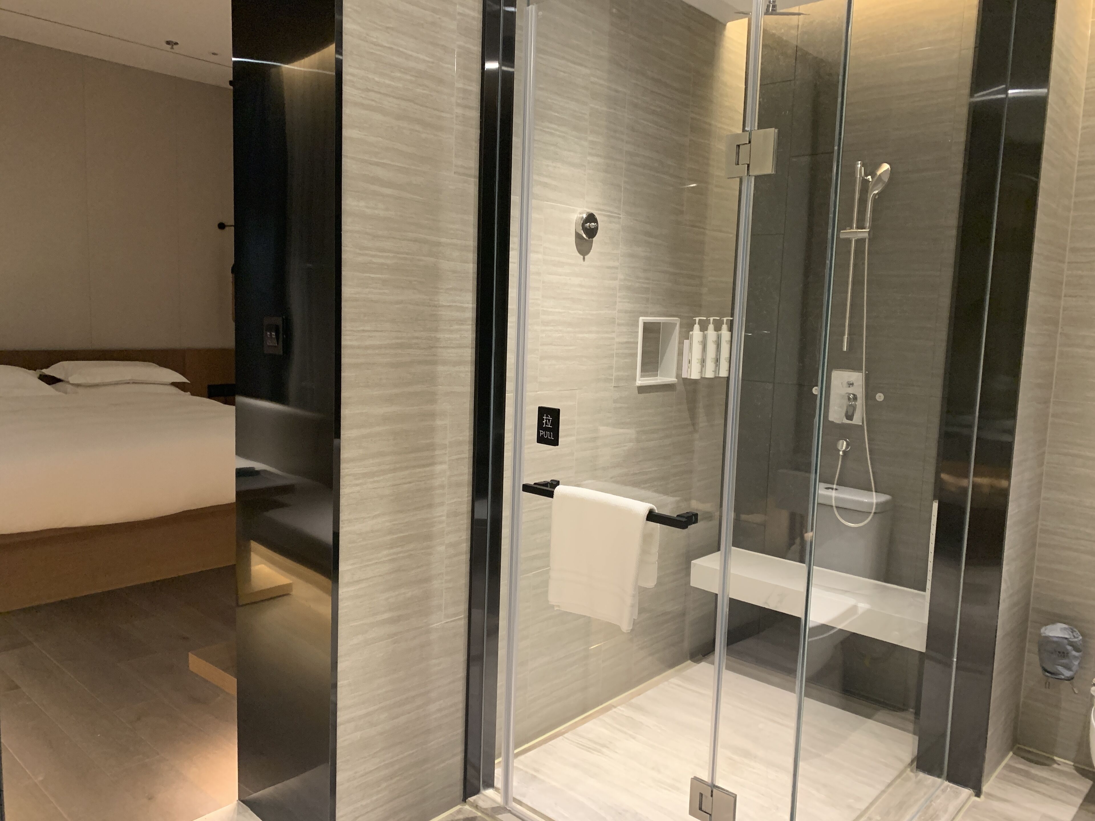 Quarto de hotel moderno com chuveiro em cabine de vidro, cama bem arrumada e pequena mesa de cabeceira com produtos de higiene pessoal.