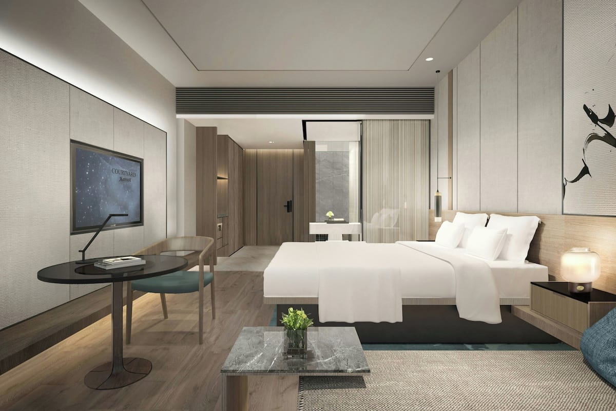 Quarto de hotel moderno com uma cama grande, uma escrivaninha com cadeira, uma mesinha de centro com um vaso e uma janela ampla com cortinas.