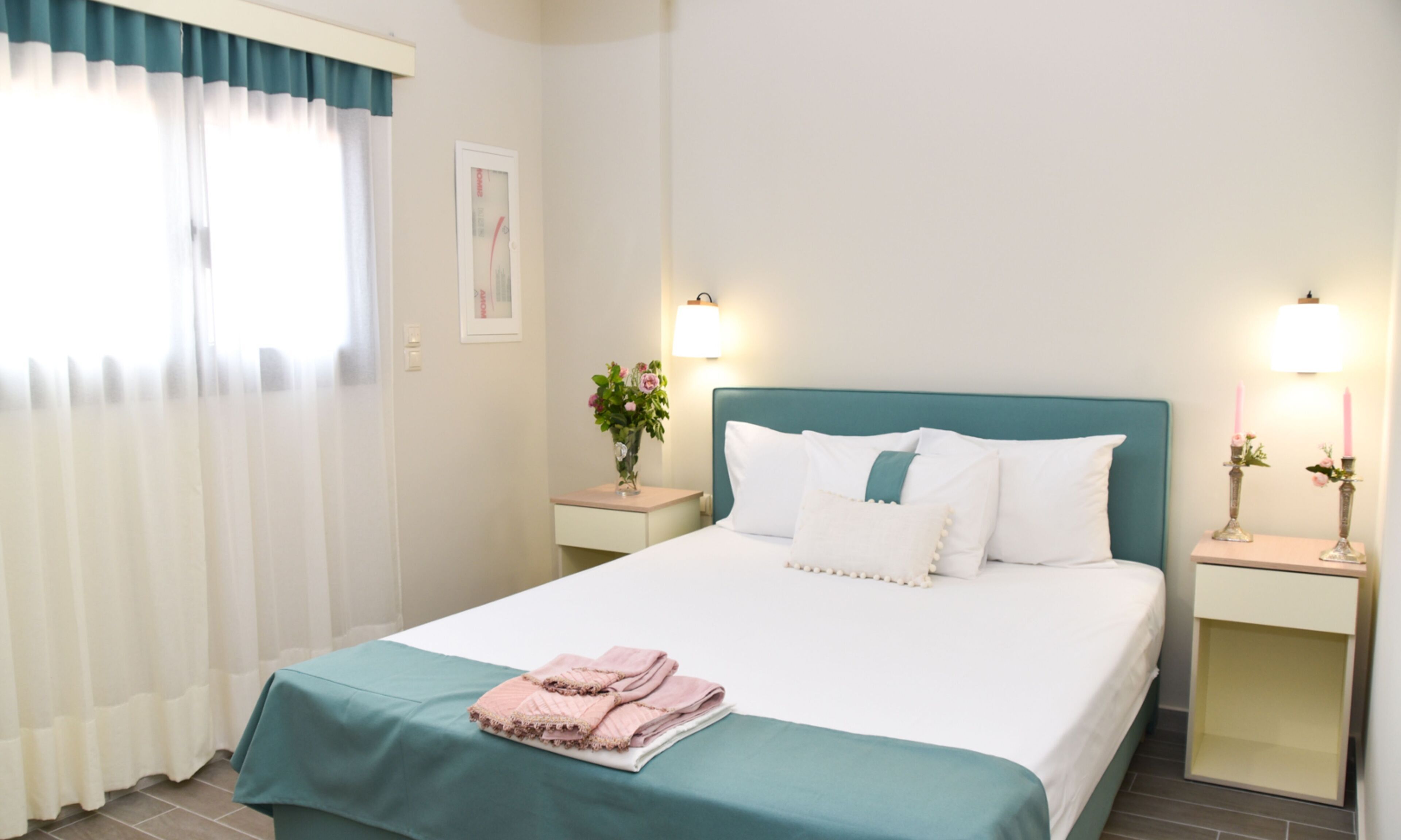 Cama bem arrumada com colcha azul-turquesa e toalhas cor-de-rosa, duas mesas de cabeceira com abajures, uma janela com cortinas translúcidas e um quadro na parede.