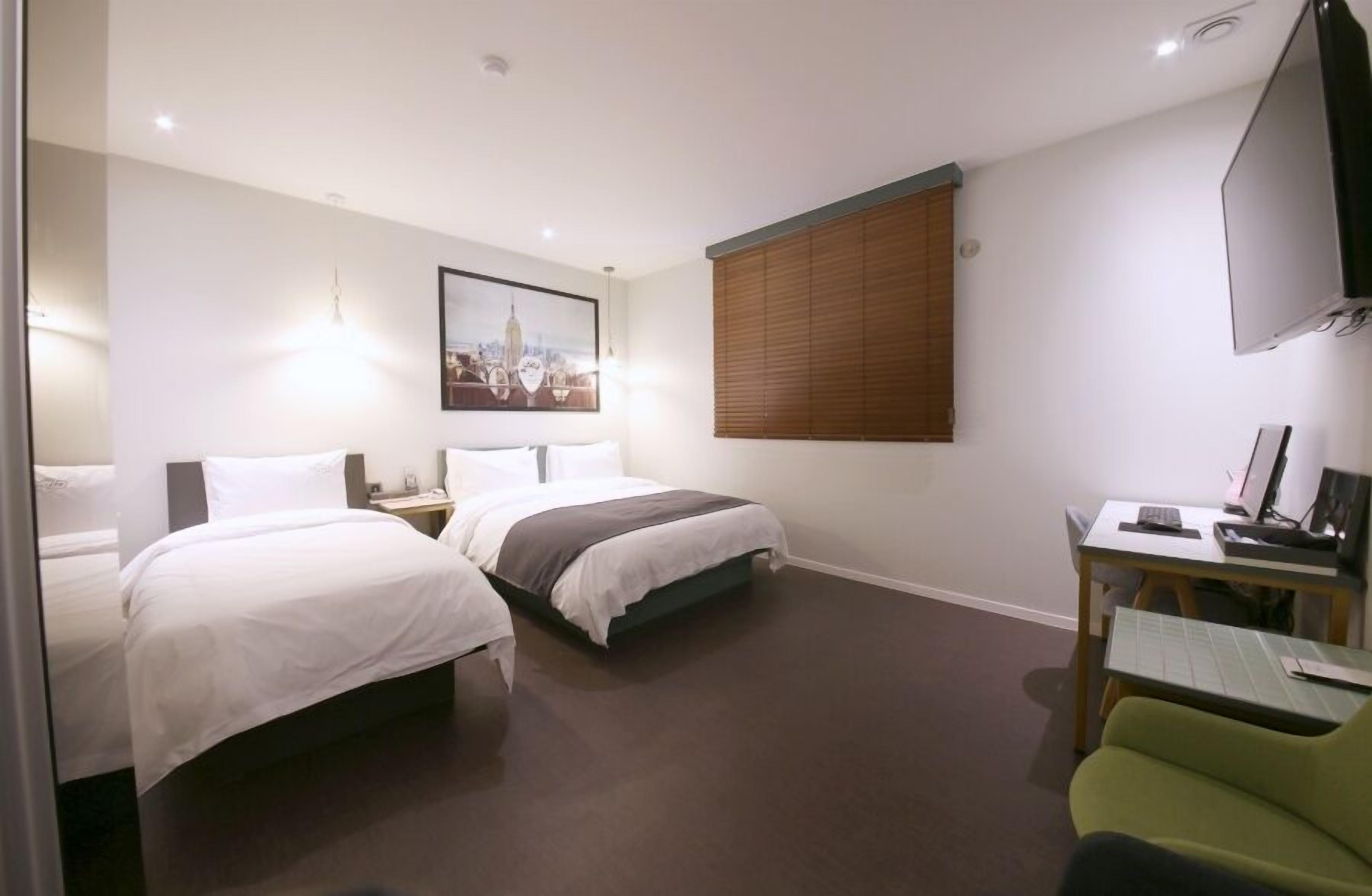 Quarto de hotel com duas camas, uma escrivaninha, uma cadeira, uma televisão e uma janela com persianas.