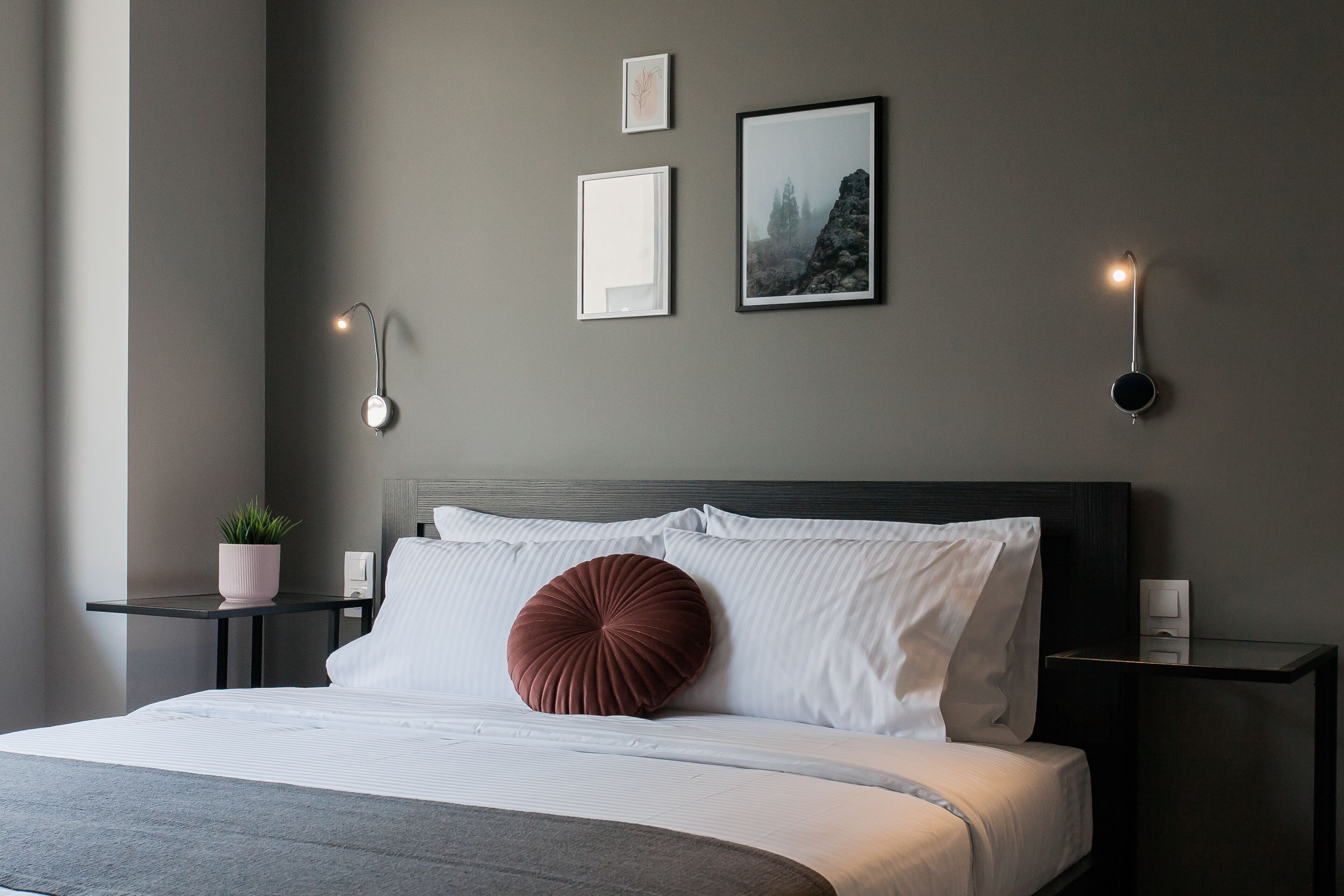 Quarto moderno com cama, mesa de cabeceira com uma planta e luminária fixa na parede.