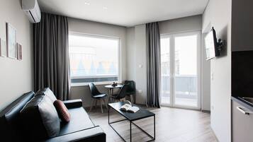 Uma sala de estar moderna com sofá, mesa de centro e TV fixada na parede. Há duas janelas com cortinas, e é visível um varanda através das janelas, além de uma pequena mesa com cadeiras.