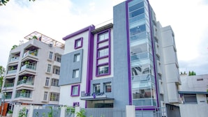 Um edifício moderno de vários andares com fachada roxa e cinzenta, varandas com plantas e entrada visível.