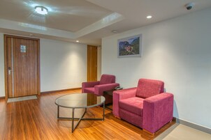 Um saguão de hotel com piso de madeira, duas poltronas cor-de-rosa, uma mesa de centro de vidro e um quadro na parede.