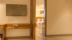 Quarto de hotel com televisão na parede, escrivaninha de madeira com tomada e cama com cabeceira branca e travesseiro roxo.