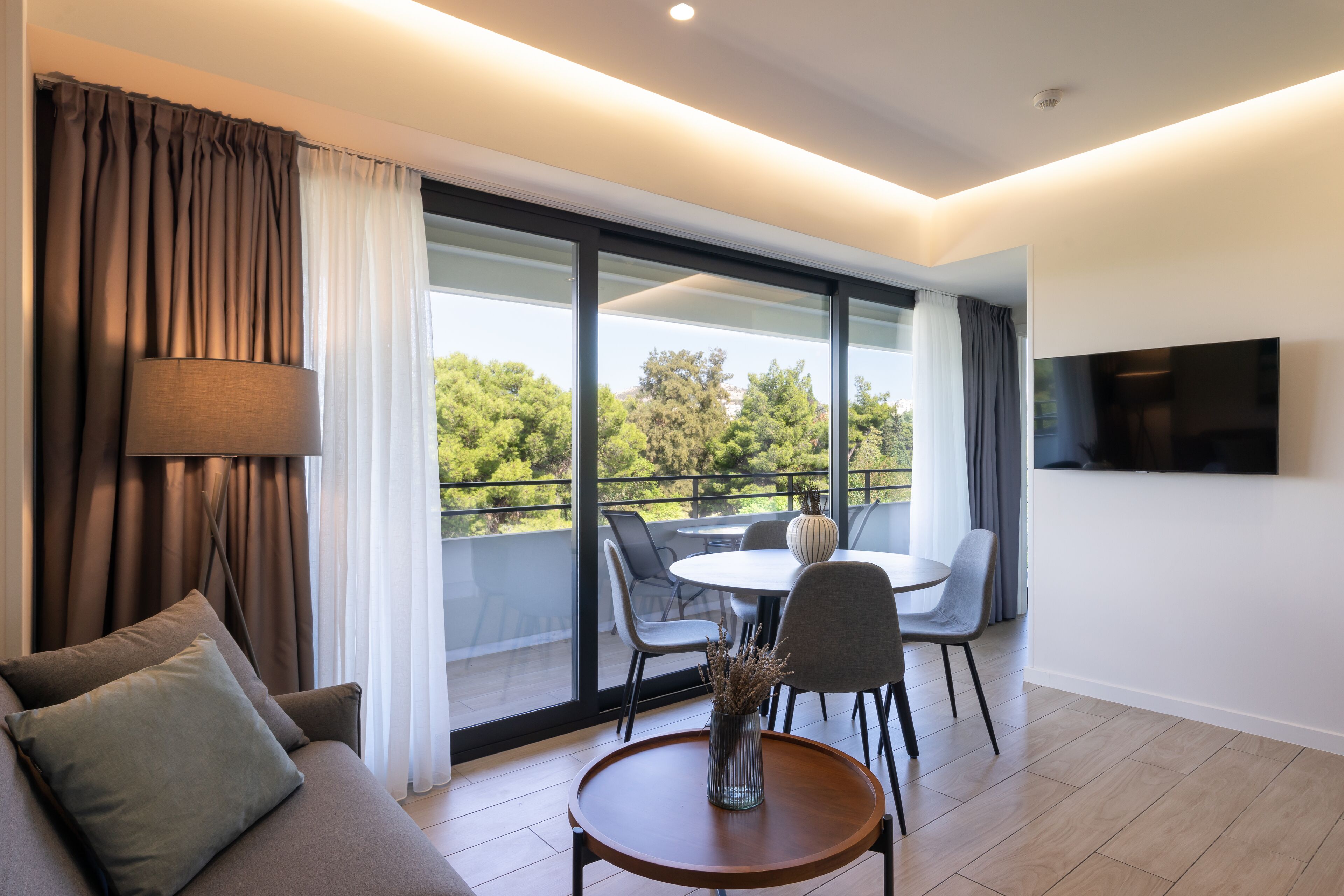 Uma sala de estar moderna com um sofá, uma mesa de centro e uma área de jantar com cadeiras e uma mesa. Uma televisão está fixada na parede e há uma janela grande com cortinas.