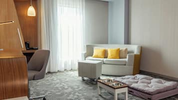 Quarto de hotel moderno com sofá cinzento, poltrona cinzenta, um banquinho pequeno, cama para animais de estimação e máquina de café sobre uma mesa.