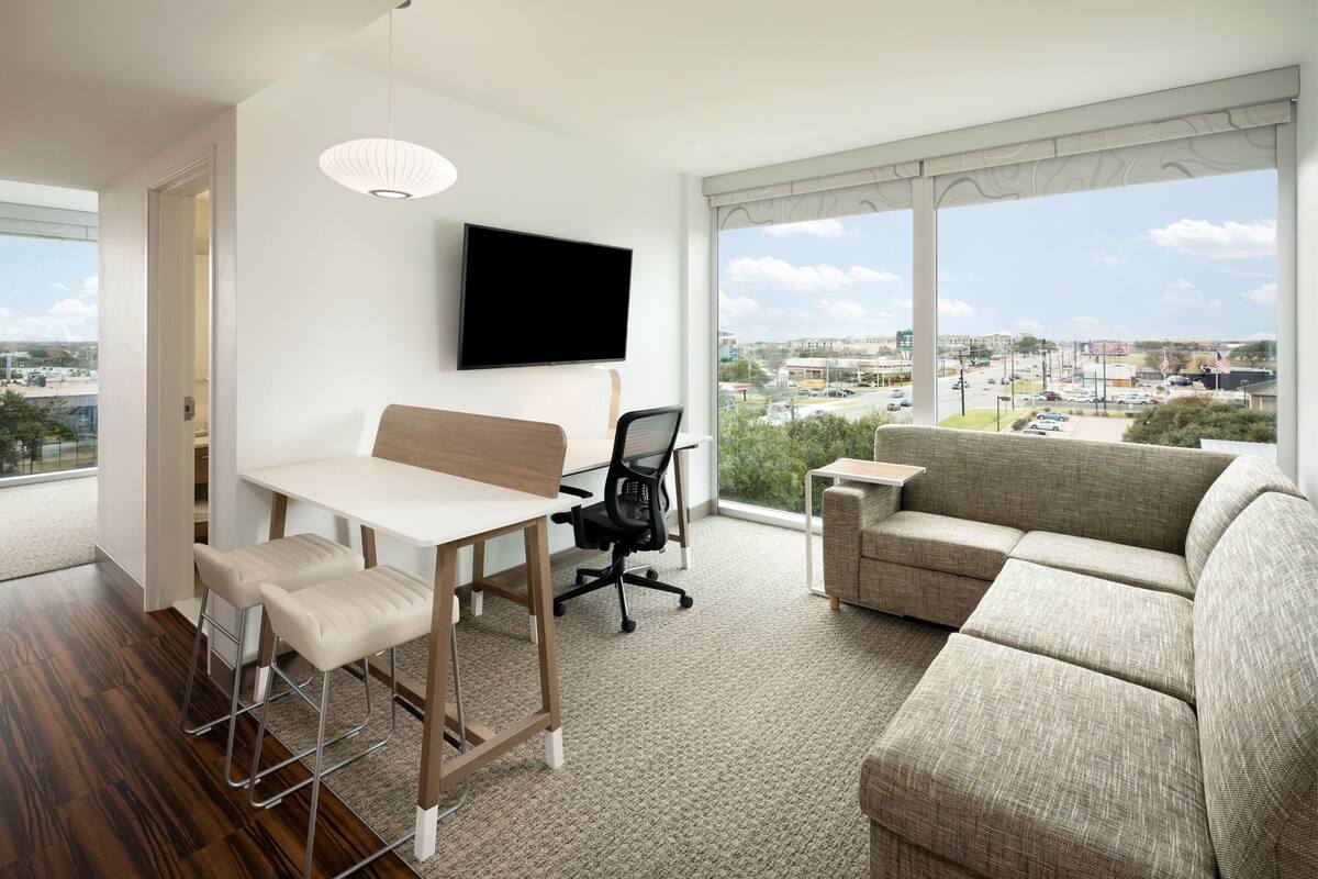 Quarto de hotel moderno com uma grande janela com vista para a cidade, televisão de tela plana na parede, escrivaninha de madeira com dois banquinhos e um sofá bege.