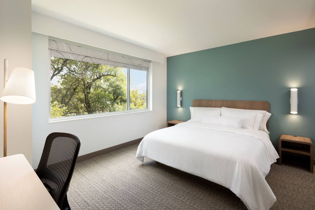 Quarto com uma cama, uma cadeira, mesa de cabeceira, abajur e janela com vista para árvores.