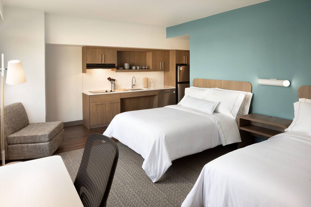Quarto de hotel com duas camas, cozinha compacta, mesa, cadeira e luminária.