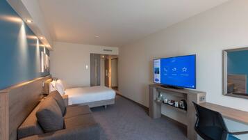 Quarto de hotel moderno com cama, sofá, escrivaninha, televisão e espelho.