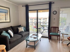 Uma sala de estar com um sofá cinza escuro, uma mesa de centro de vidro e vista para uma varanda com mobiliário externo.