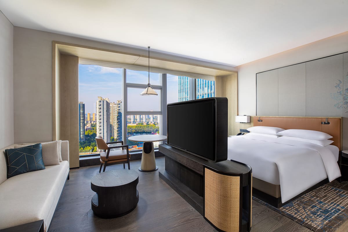 Quarto de hotel moderno com uma cama grande, televisão, sofá, mesa de jantar e vista para a cidade.