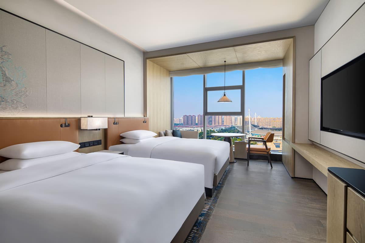Quarto de hotel com duas camas, uma escrivaninha, televisão e uma janela grande com vista para a cidade.