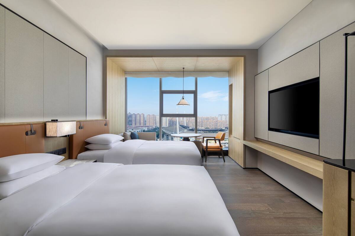 Quarto de hotel com duas camas, televisão, vista para a cidade e área de refeições.