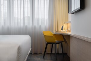 Quarto de hotel com cama, escrivaninha com cadeira, televisão de tela plana e janela com cortinas leves.