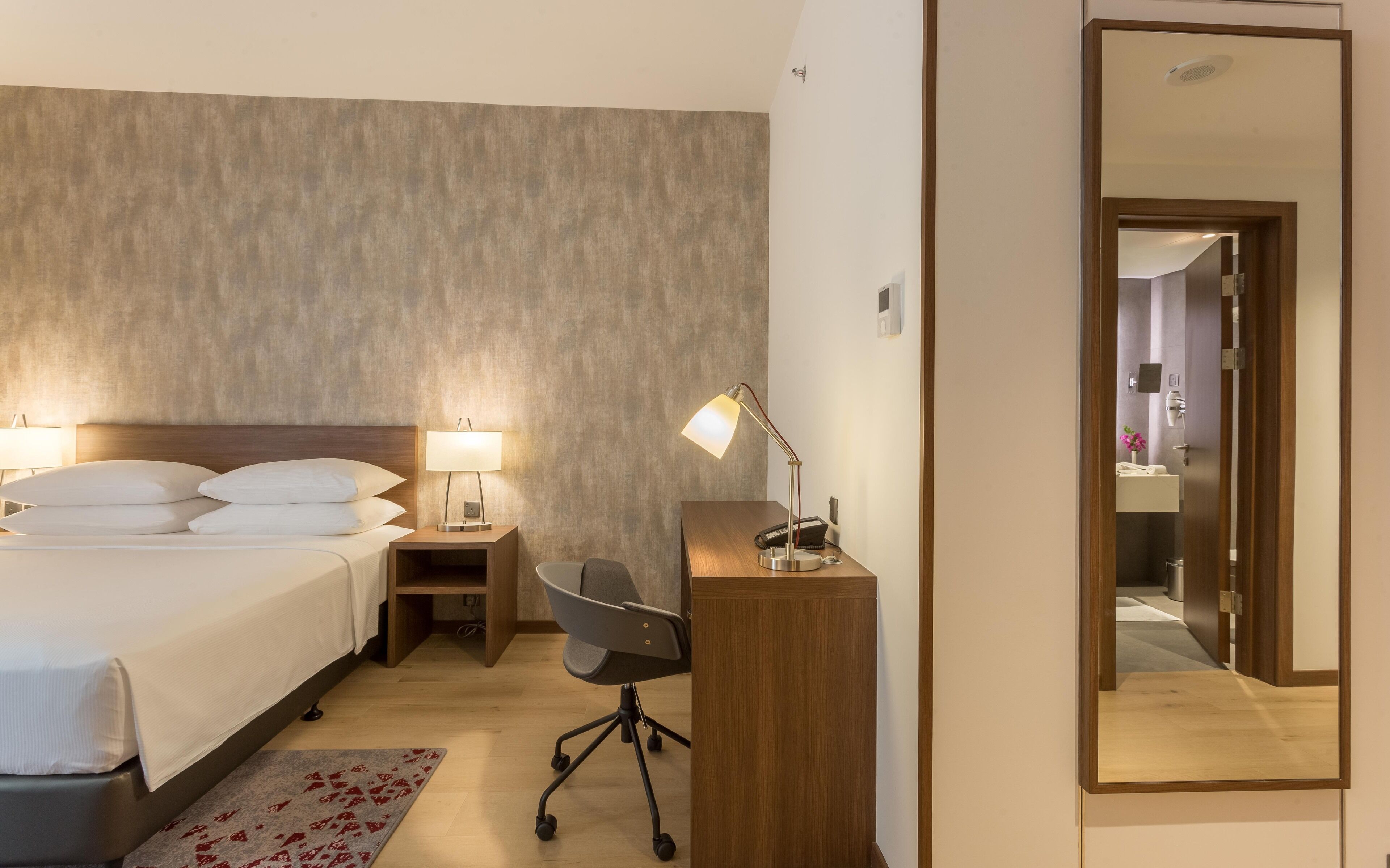 Quarto de hotel com uma cama grande, uma escrivaninha com luminária, uma cadeira e um espelho.