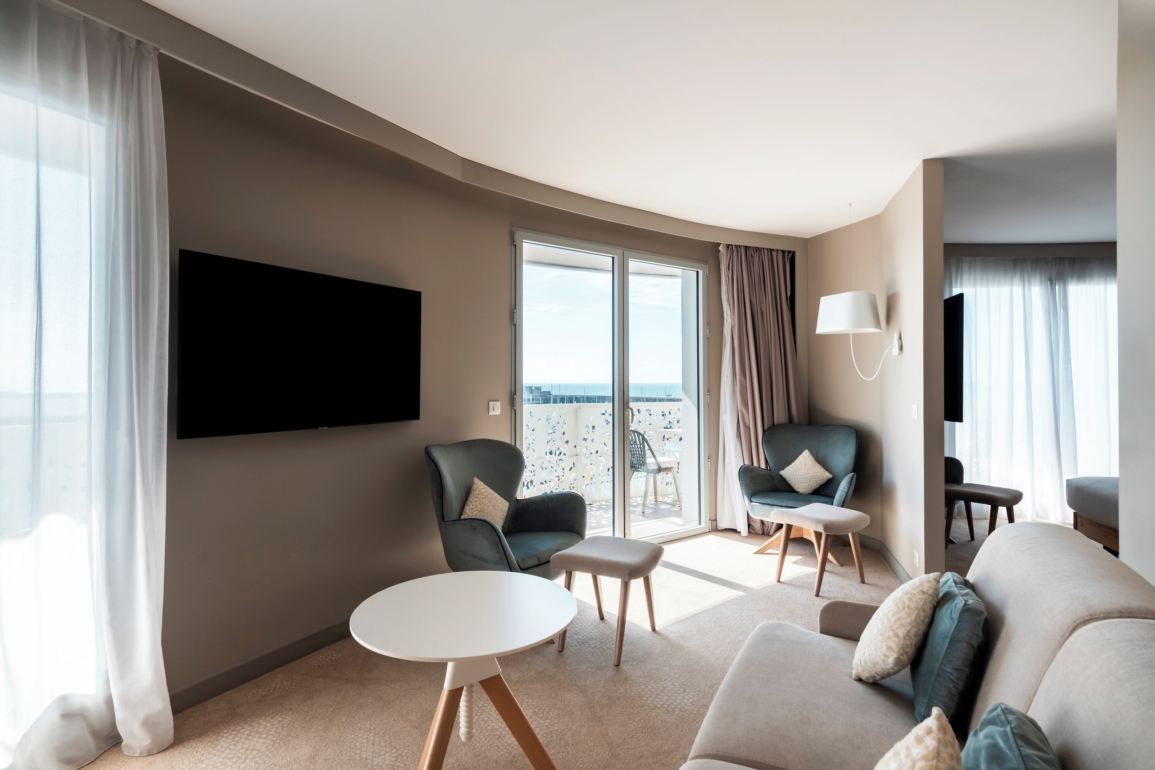 Quarto de hotel moderno com teto curvo, TV de tela plana na parede, área de estar com sofá e cadeiras, mesa redonda pequena e varanda com vista.