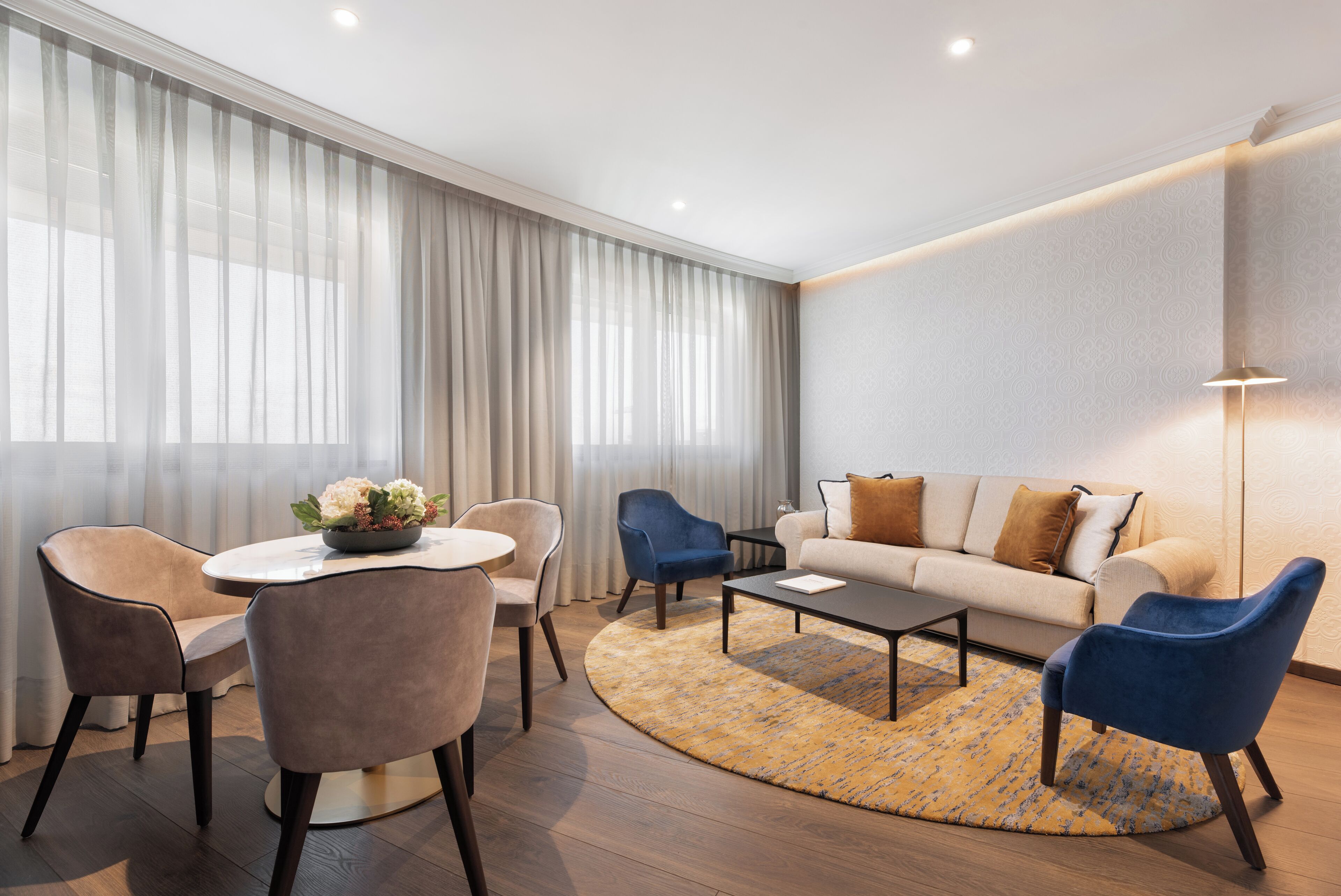 Uma sala de estar moderna com uma mesa de centro redonda, sofá bege, poltronas azuis e uma área de refeições com mesa e cadeiras.