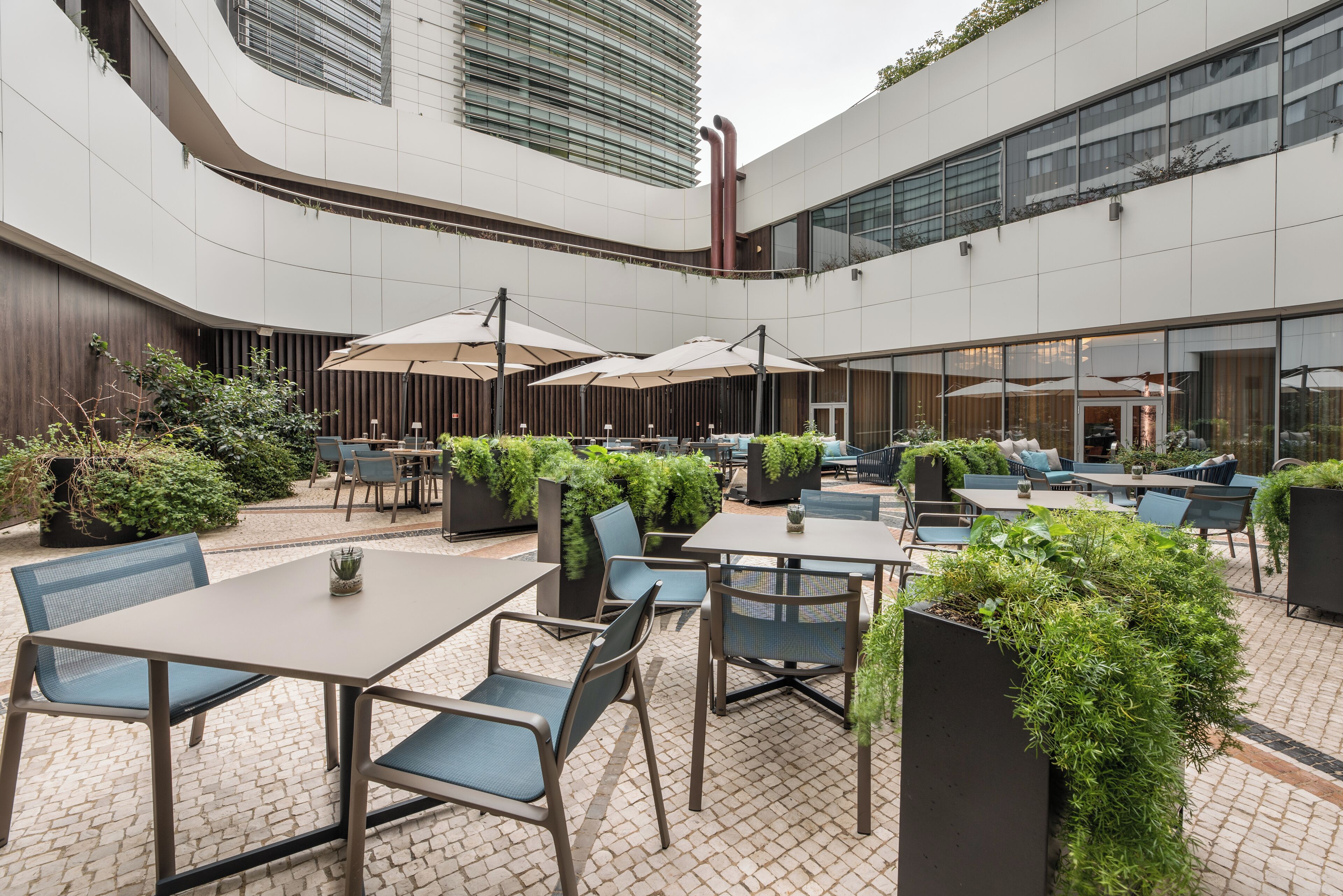 Área de terraço ao ar livre com mesas e cadeiras, rodeada por vegetação e arquitetura moderna.