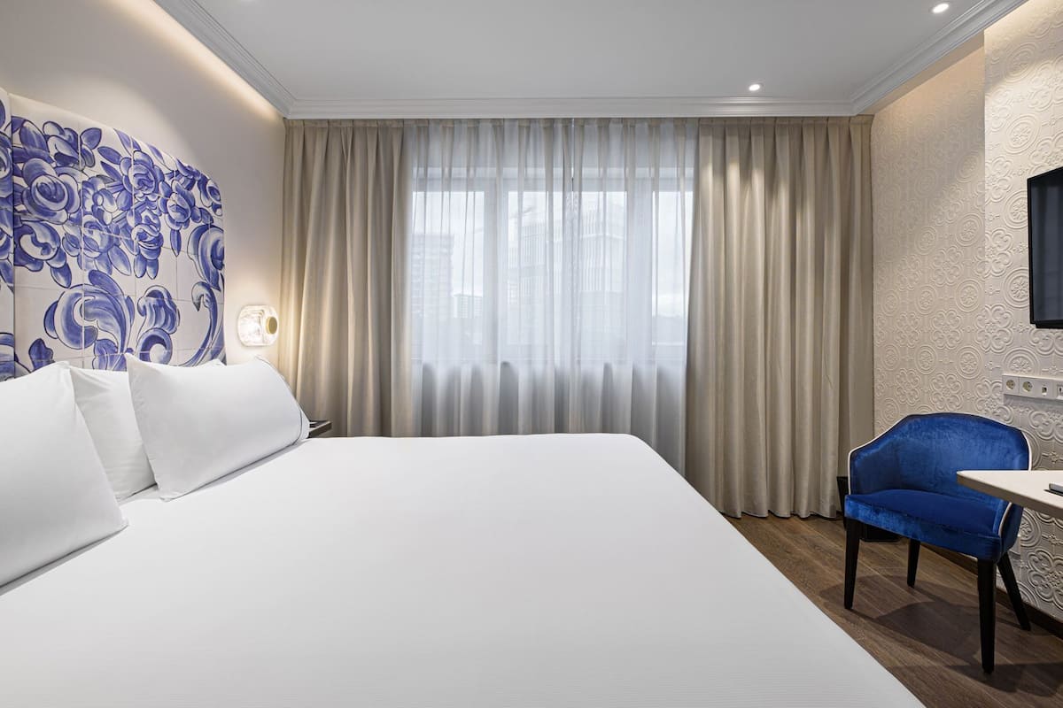 Quarto de hotel com uma cama grande, uma escrivaninha, uma cadeira azul e uma televisão na parede.