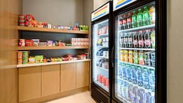 Uma área de exposição de lanches e bebidas, com prateleiras repletas de diversos petiscos e uma geladeira cheia de água engarrafada e refrigerantes.