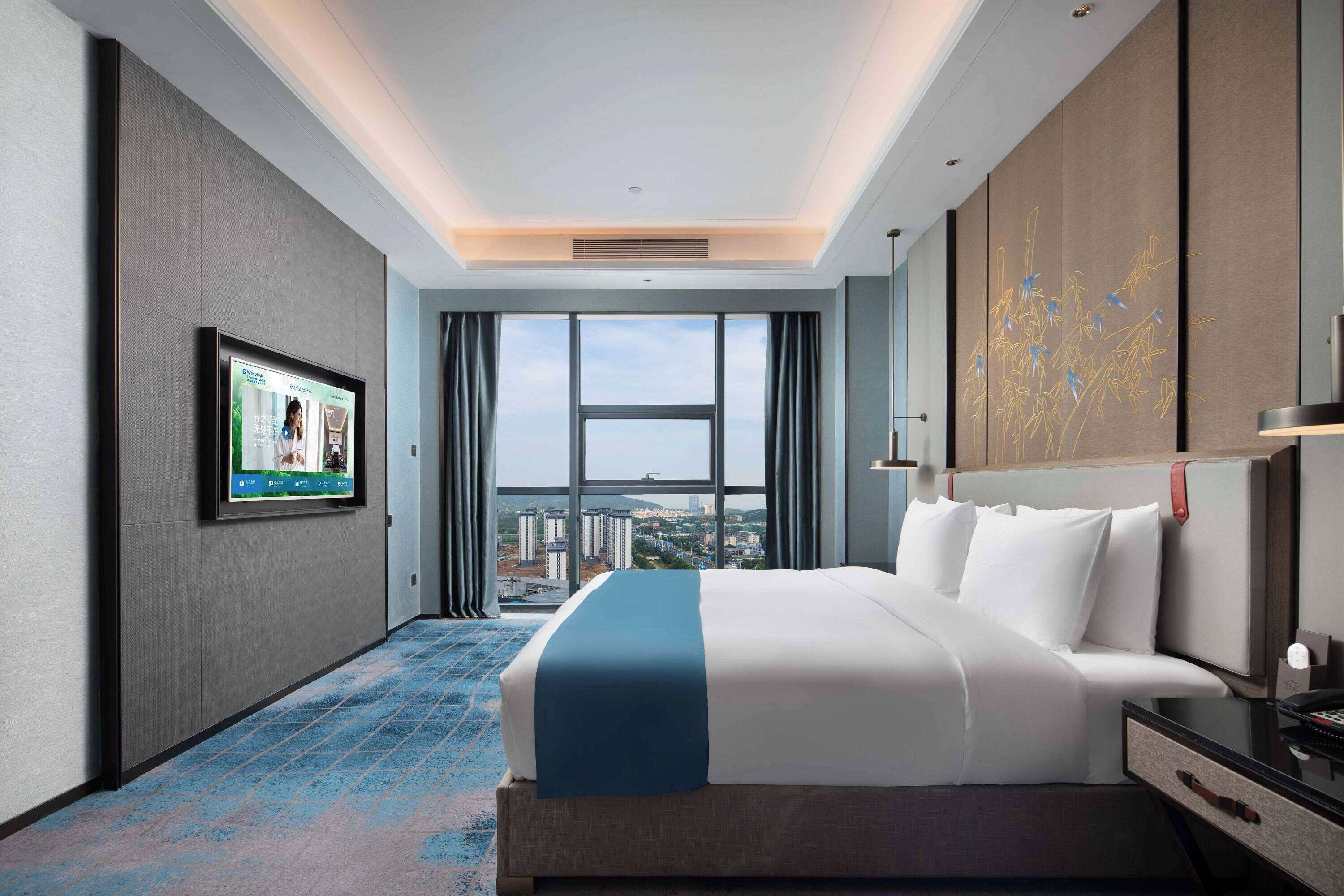 Quarto de hotel moderno com uma cama grande, televisão na parede e vista da cidade através de amplas janelas.
